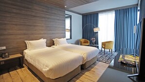 Quarto de hotel com duas camas, uma escrivaninha com luminária, uma cadeira e uma janela grande com cortinas.