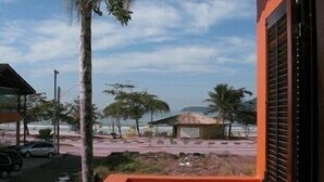 Um edifício com fachada vermelha e varanda com vista para uma rua com palmeiras e céu claro.