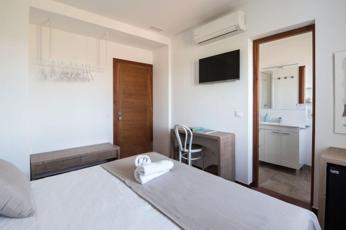 Quarto de hotel com cama, escrivaninha, cadeira, guarda-roupa, televisão e banheiro visível por uma porta aberta.
