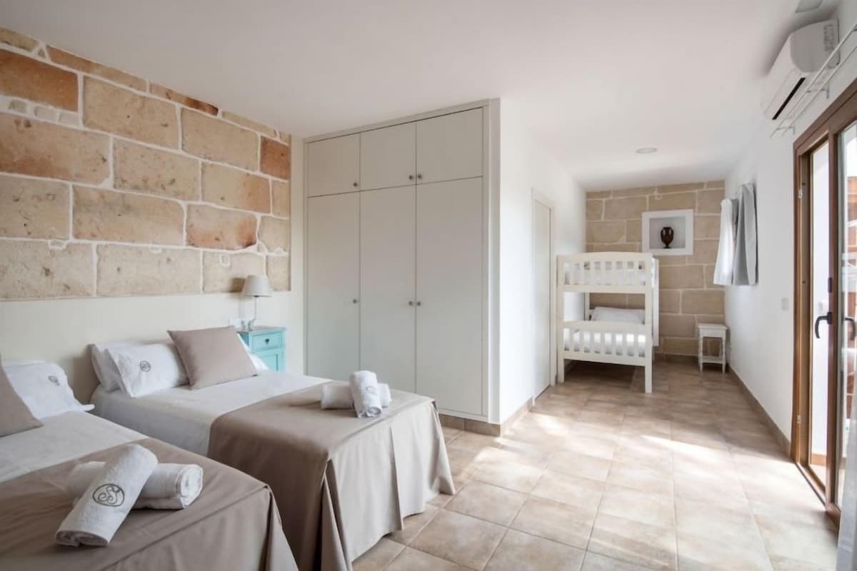 Quarto de hotel com duas camas, um guarda-roupa, uma mesa pequena e um banco.