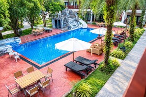 Piscina com espreguiçadeiras e mesas, cercada por vegetação e um elemento decorativo de pedras.