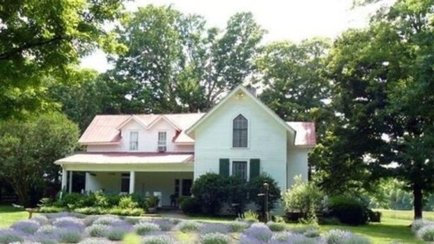 Uma casa de dois andares com telhado triangular, rodeada por um jardim bem cuidado com plantas de lavanda.