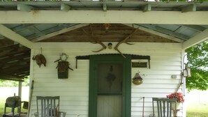 Varanda com duas cadeiras de madeira, uma porta verde e várias decorações, incluindo chifres de veado e uma placa de boas-vindas.