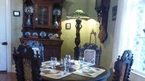 Uma sala de jantar com uma mesa redonda posta para quatro pessoas, rodeada por cadeiras elaboradas. Um armário de madeira com louças é visível ao fundo.