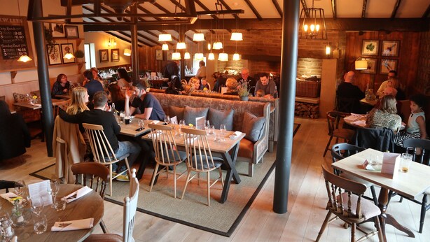 Um restaurante com mesas e bancos de madeira, luminárias pendentes e uma parede de tijolos.