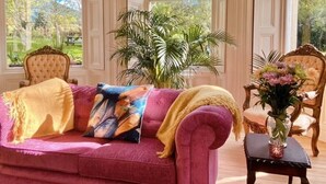 Quarto com um sofá rosa, uma mesinha com um vaso e janelas grandes com vista para árvores.