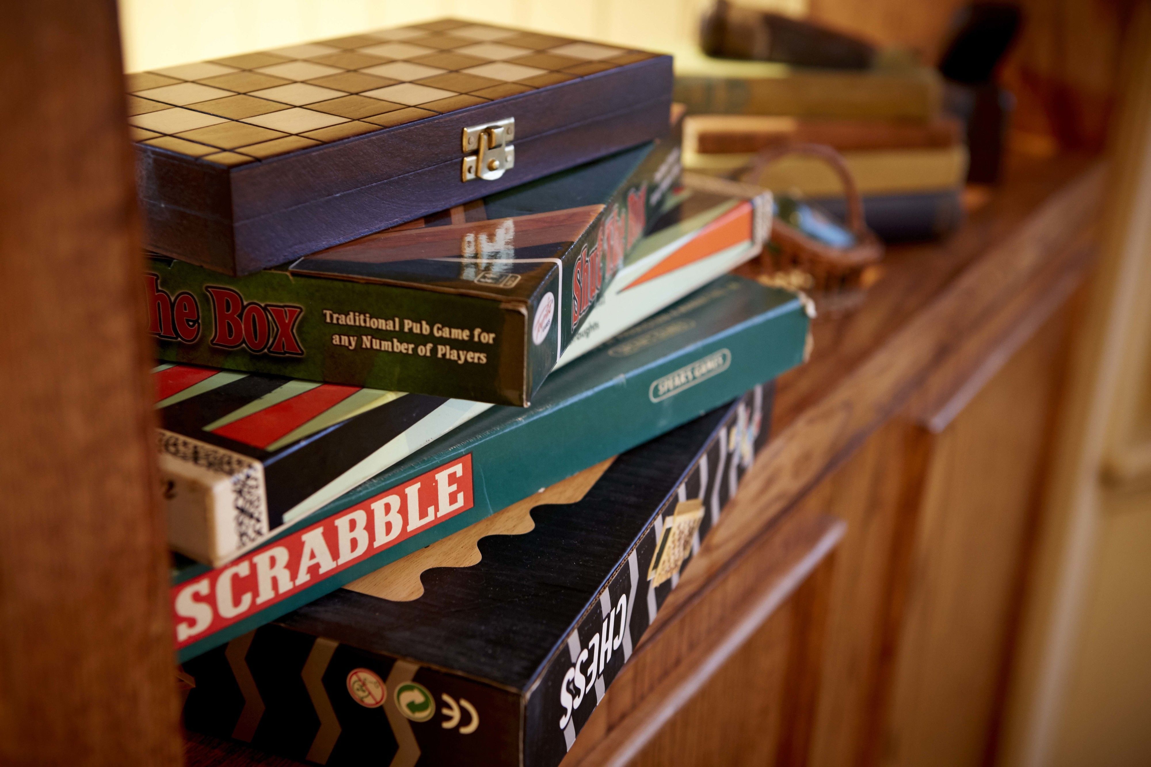 Uma pilha de jogos de tabuleiro, incluindo Scrabble e The Box, sobre uma superfície de madeira.