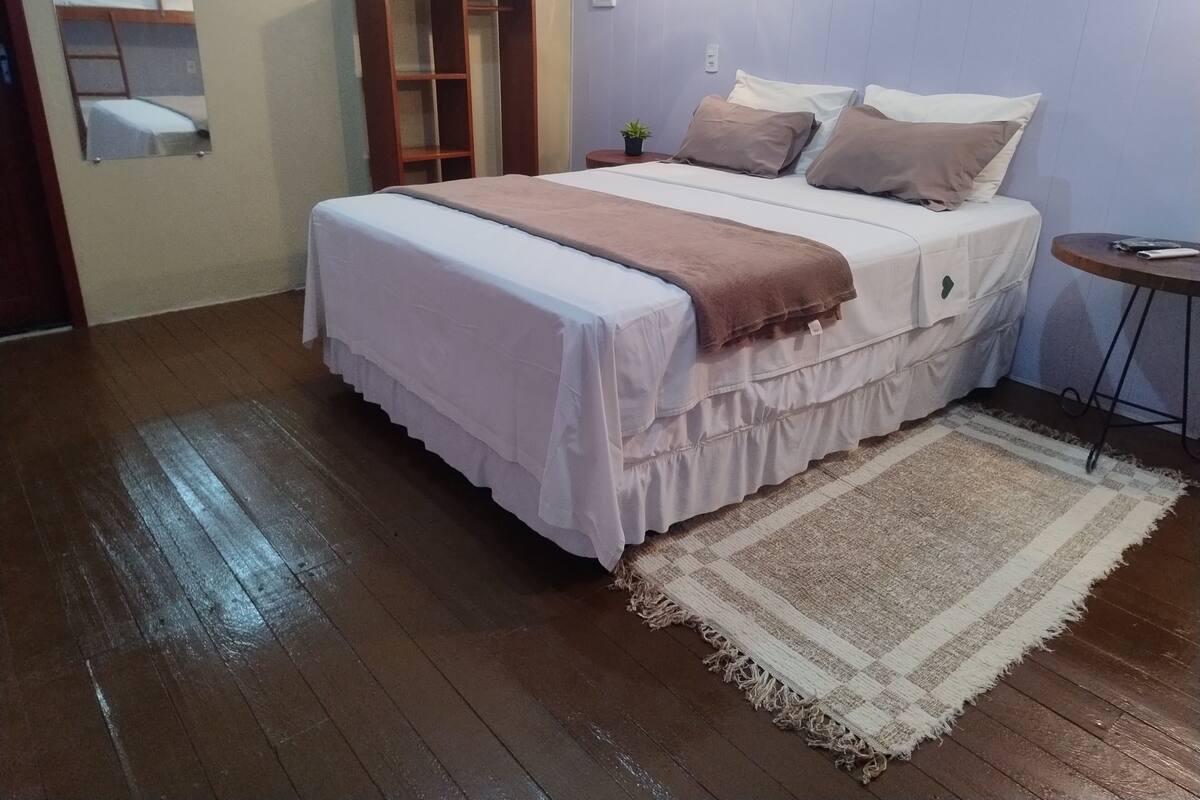 Quarto com cama, guarda-roupa de madeira, mesa pequena e prateleira na parede.