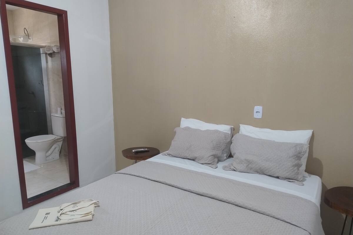 Quarto de hotel com cama, duas almofadas, mesa de cabeceira, espelho e casa de banho visível através de uma porta.