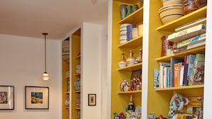 Quarto com piso de madeira, uma estante cheia de livros e objetos decorativos, duas cadeiras e uma mesa com diversos objetos.