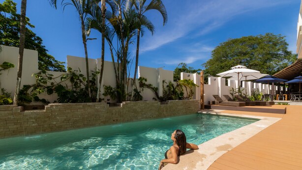 Uma área de piscina com água cristalina, deck de madeira e palmeiras.