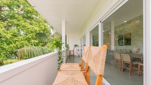 Varanda com cadeiras de vime, espaço de jantar com cadeiras e mesa de madeira, e vista para vegetação exuberante.