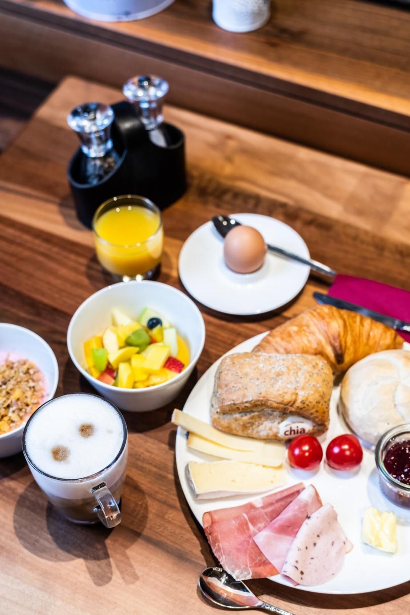 Uma mesa de madeira com um pequeno-almoço que inclui croissant, fiambre, queijo, ovos, fruta e bebidas.