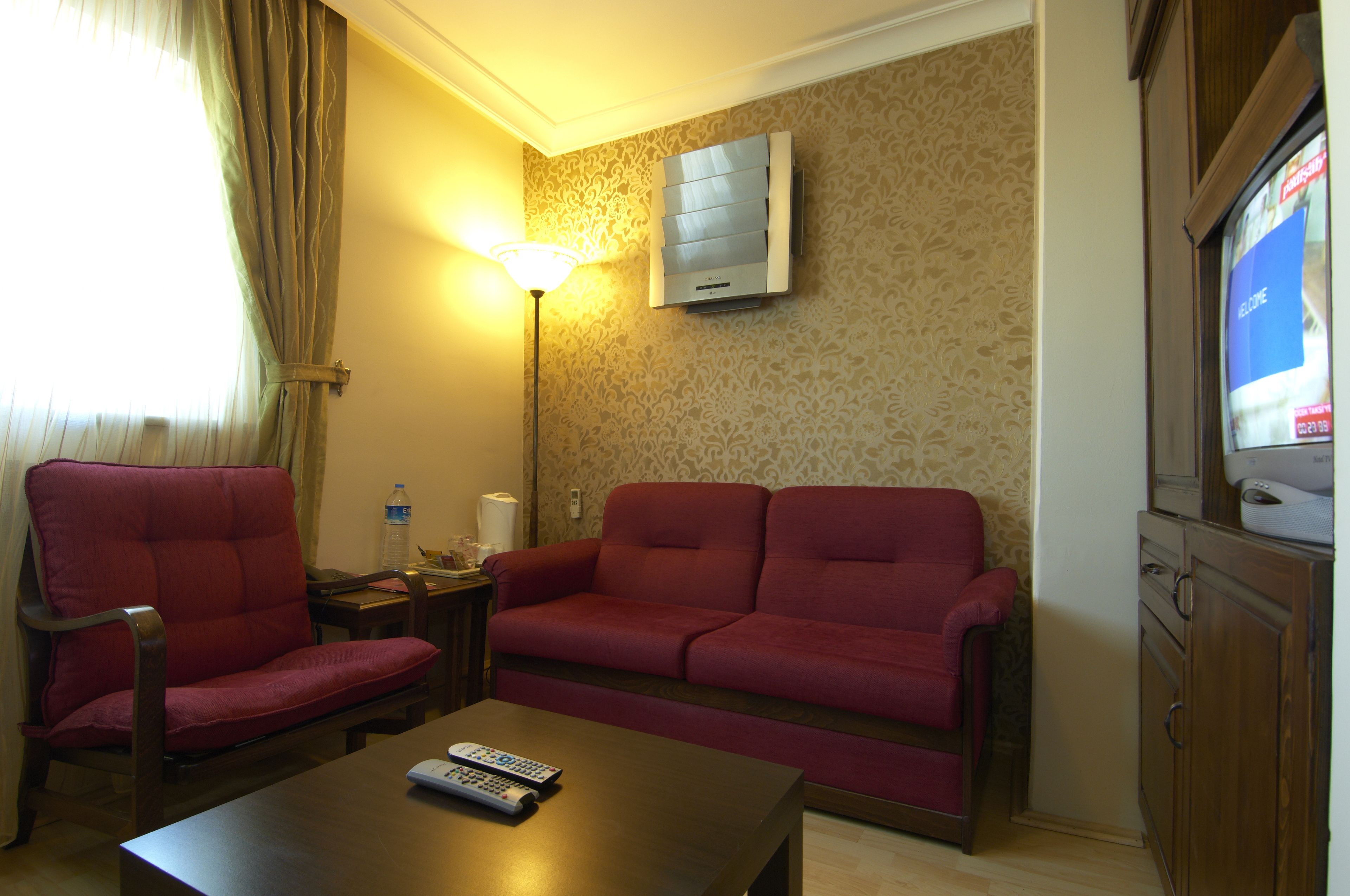 Quarto de hotel com um sofá vermelho, uma mesa de centro de madeira com um controle remoto, uma luminária e uma televisão na parede.