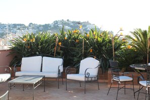 Uma área externa com assentos, incluindo cadeiras brancas acolchoadas e uma mesa de metal, cercada por plantas verdes e vista para uma cidade no alto de uma colina.
