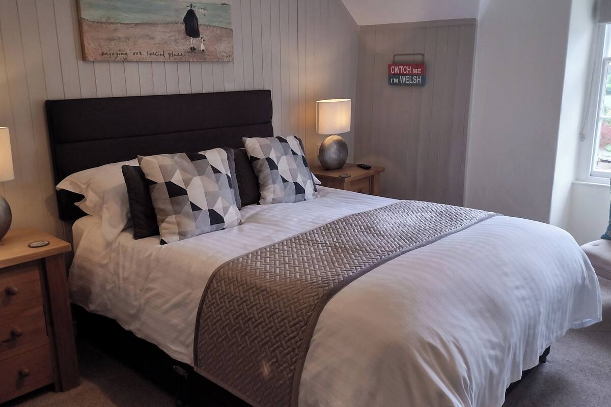Cama bem arrumada com colcha estampada e almofadas, duas luminárias de cabeceira, mesa de cabeceira de madeira, uma pequena gaveta e uma placa na parede.