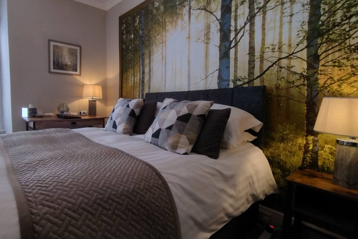Quarto de hotel com uma cama grande, mesa de cabeceira com abajur, quadro de uma floresta na parede e janela com vista para árvores.