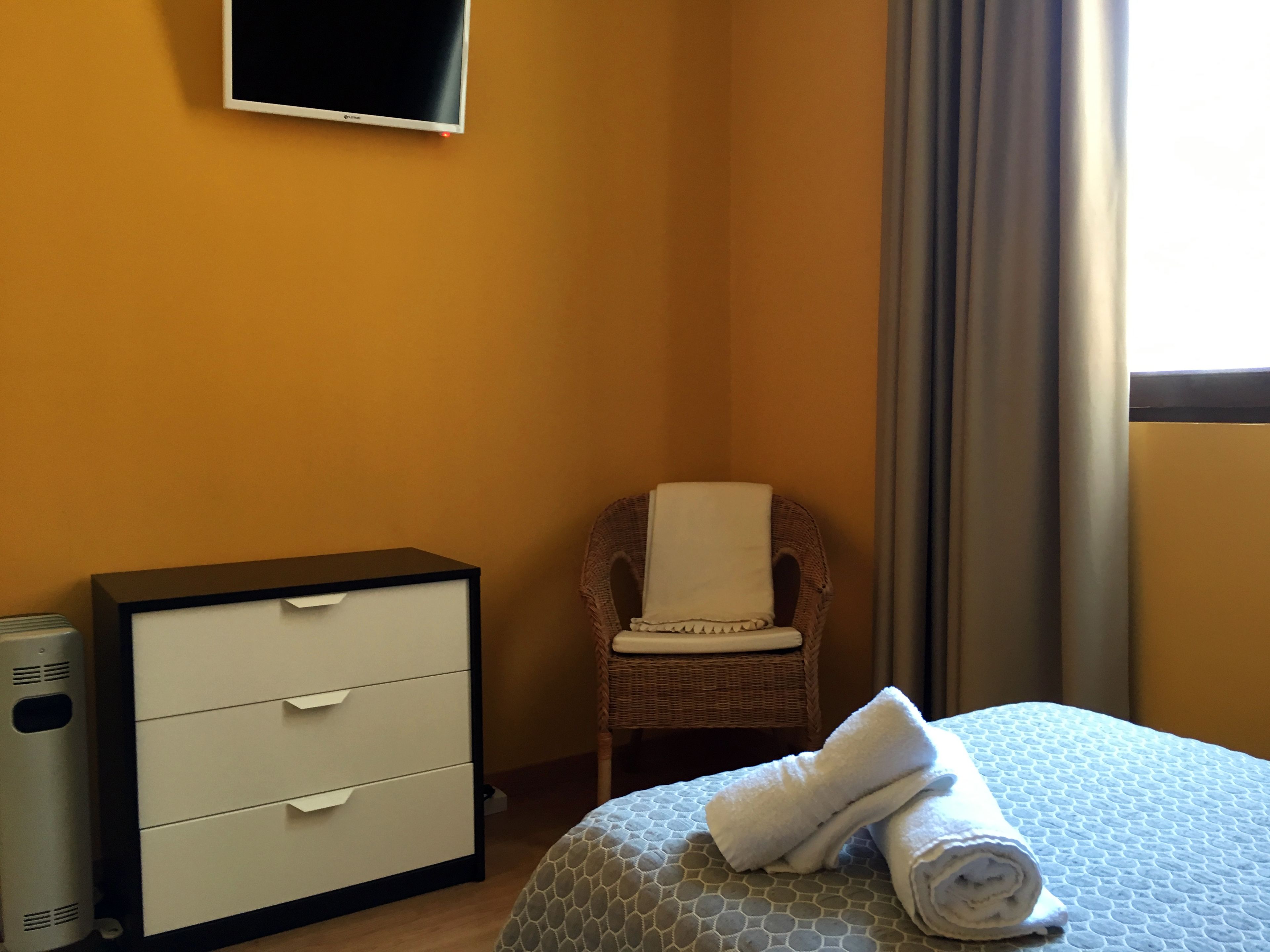 Quarto de hotel com cama, cadeira, escrivaninha e janela com cortinas.