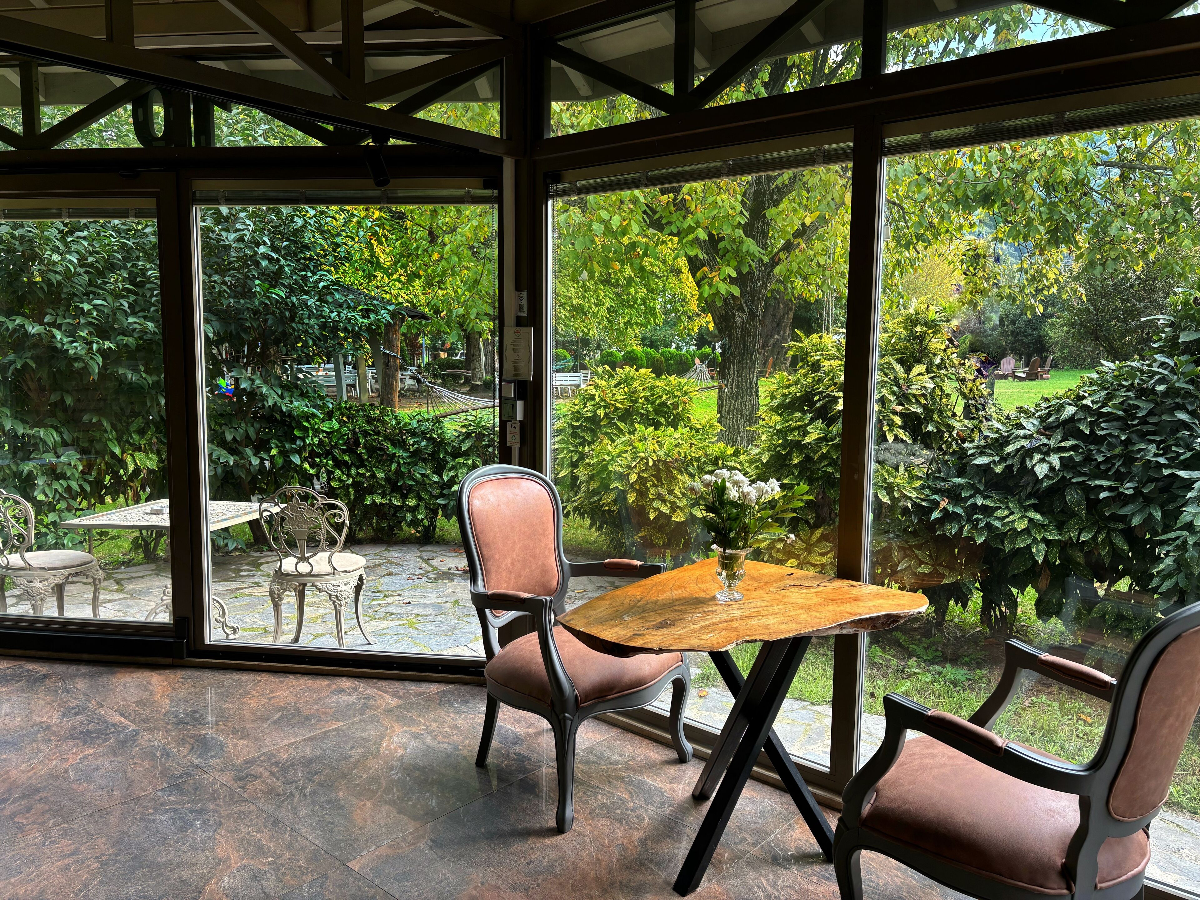 Uma estufa com mesa e cadeiras, amplas janelas e vista para um jardim.