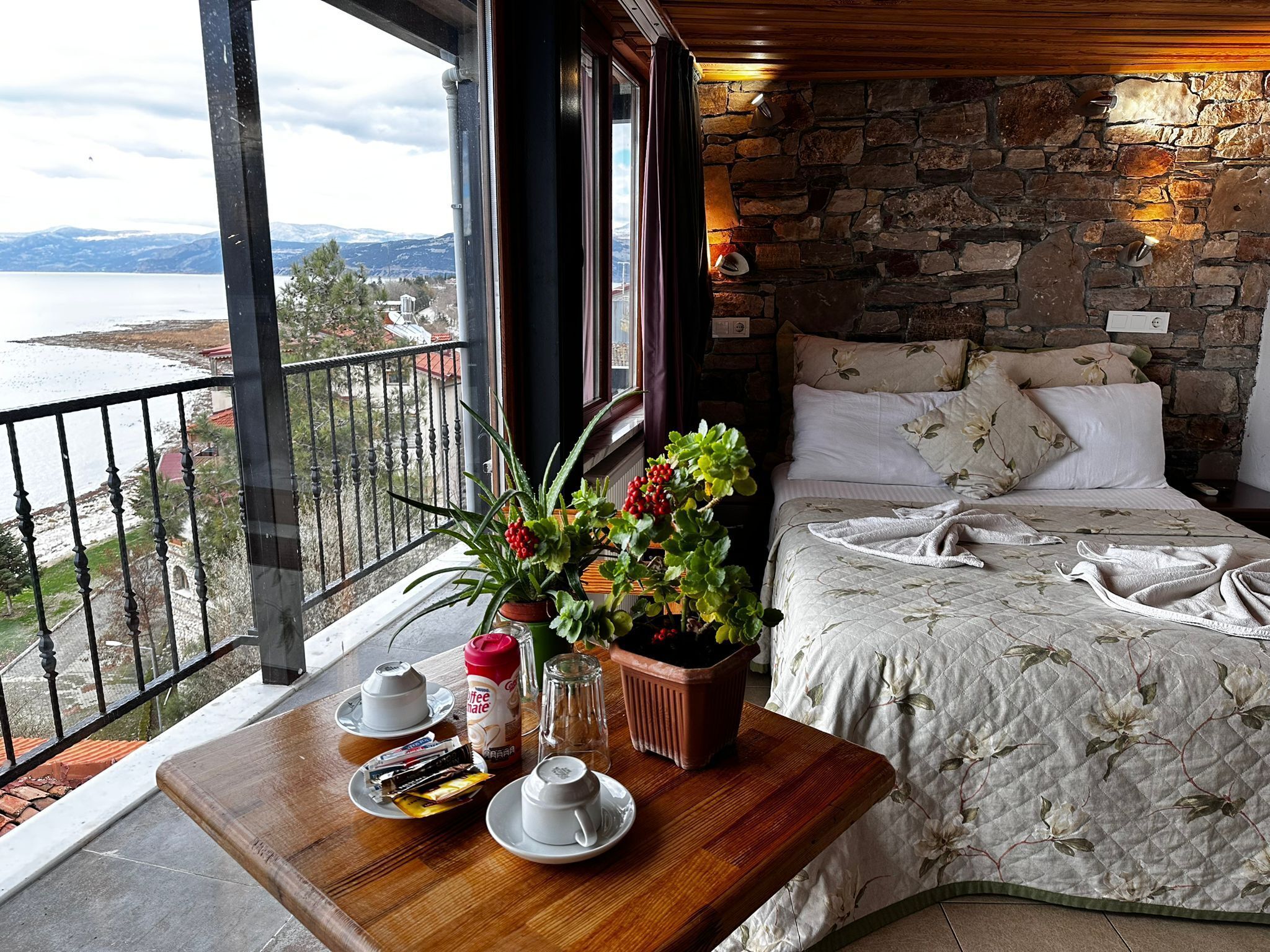 Uma varanda com uma mesa de madeira, uma cama com roupa de cama florida e uma parede de pedra.