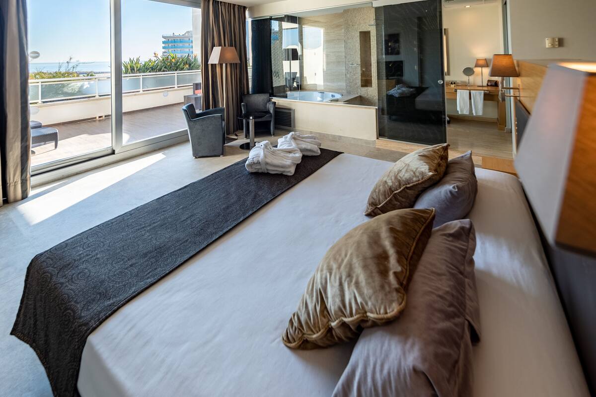 Quarto de hotel moderno com uma cama grande, um espaço de estar com uma poltrona, uma pequena escrivaninha e vista para o mar.