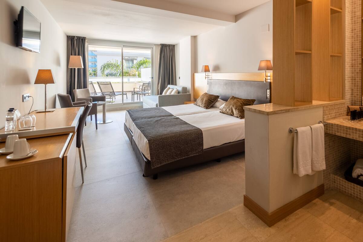 Quarto de hotel com cama, escrivaninha, poltrona, sofá, varanda e banheiro com pia e espelho.