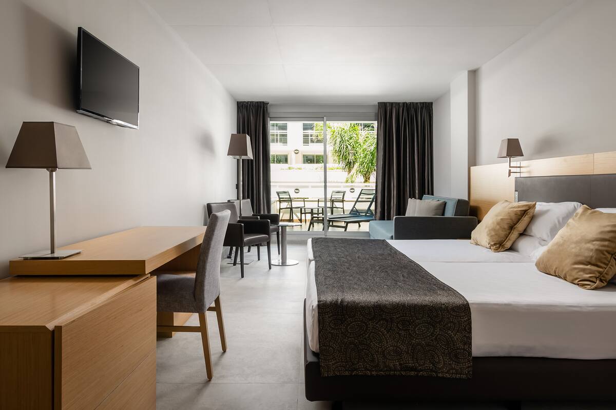 Quarto de hotel com cama, escrivaninha e televisão. Possui uma varanda com mesa e cadeiras.