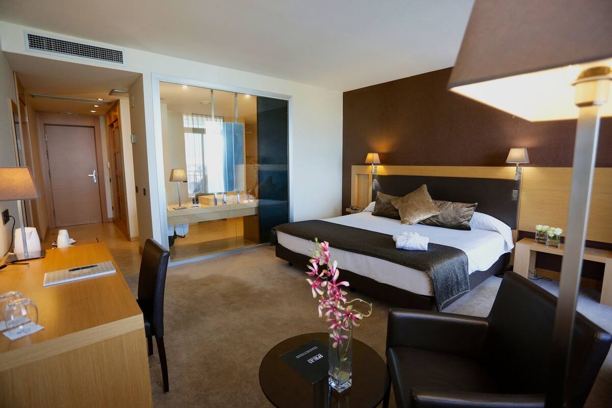 Quarto de hotel com cama, abajures, escrivaninha com cadeira, pequena mesa com um vaso de flores e um espelho.