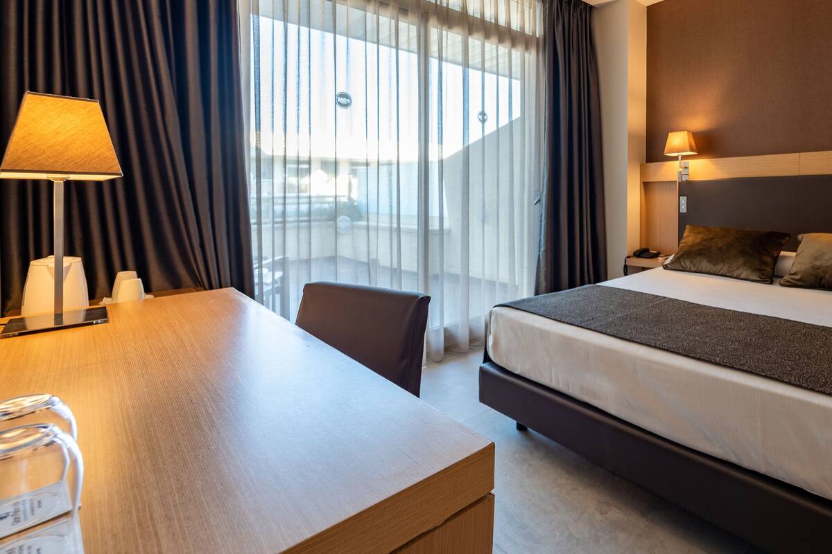 Quarto de hotel com uma cama grande, abajures, uma escrivaninha com luminária e vista através de cortinas finas.