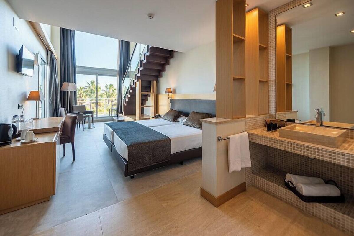 Quarto de hotel moderno com uma cama grande, uma mesa de escritório, banheiro com um espelho grande e uma escada que leva a uma área externa.