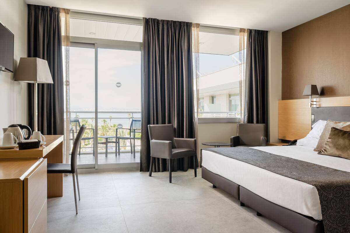 Quarto de hotel com cama, escrivaninha, cadeiras e varanda com cortinas.