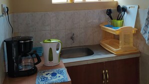 Uma cozinha compacta com janela, pia, cafeteira, chaleira e uma pequena prateleira com louças.