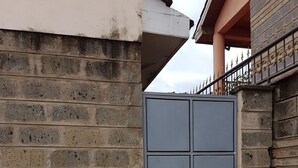 Um portão de metal cinzento com uma pequena janela circular, fixado numa base de concreto, em frente a uma parede de tijolos.