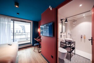 Quarto de hotel moderno com uma janela grande, uma escrivaninha, uma televisão de tela plana, um banheiro com pia e chuveiro, e uma parede de destaque vermelha.