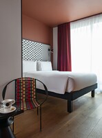 Quarto de hotel com cama, uma cadeira, uma mesa com xícara e pires, e cabeceira estampada.
