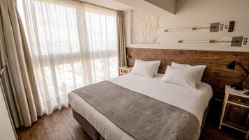 Quarto com uma cama grande, cabeceira de madeira, mesa de cabeceira com abajur e vista para o exterior através de cortinas leves.