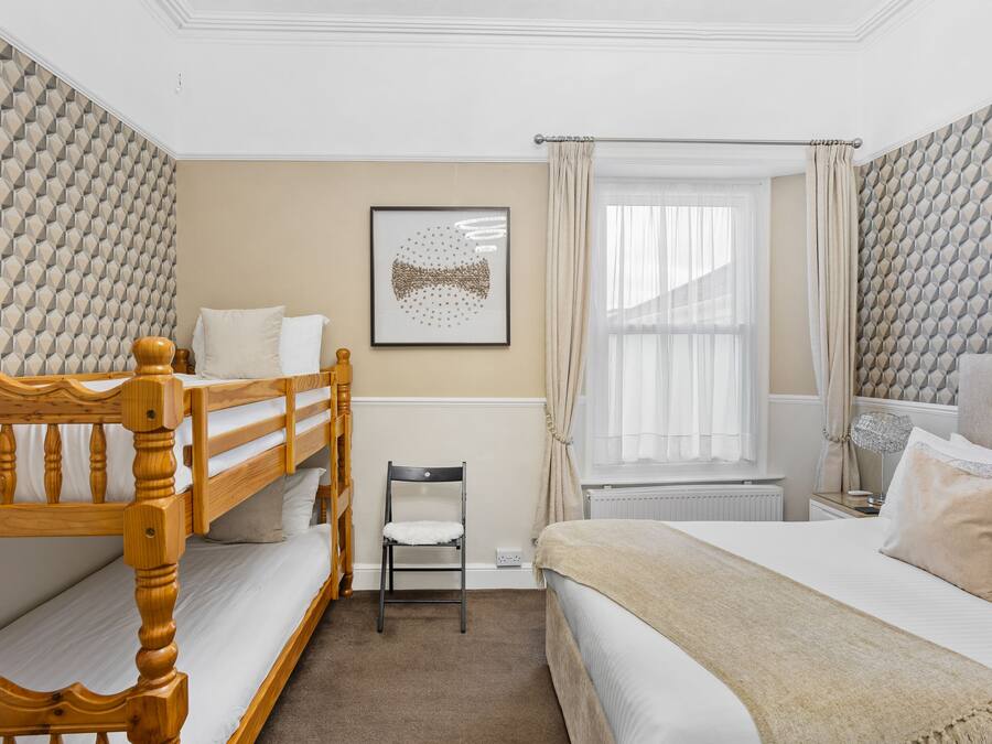 Quarto com cama beliche, cama de solteiro, cadeira, janela com cortinas e um quadro na parede.