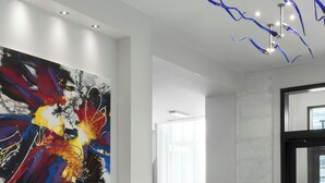 Um interior moderno com piso de mármore, uma mesa redonda de madeira com pés de metal, um quadro abstrato colorido na parede e um teto com luminárias azuis.