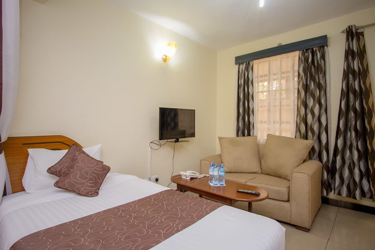 Quarto de hotel com cama, sofá, televisão, mesa de cabeceira com controle remoto e cortinas.