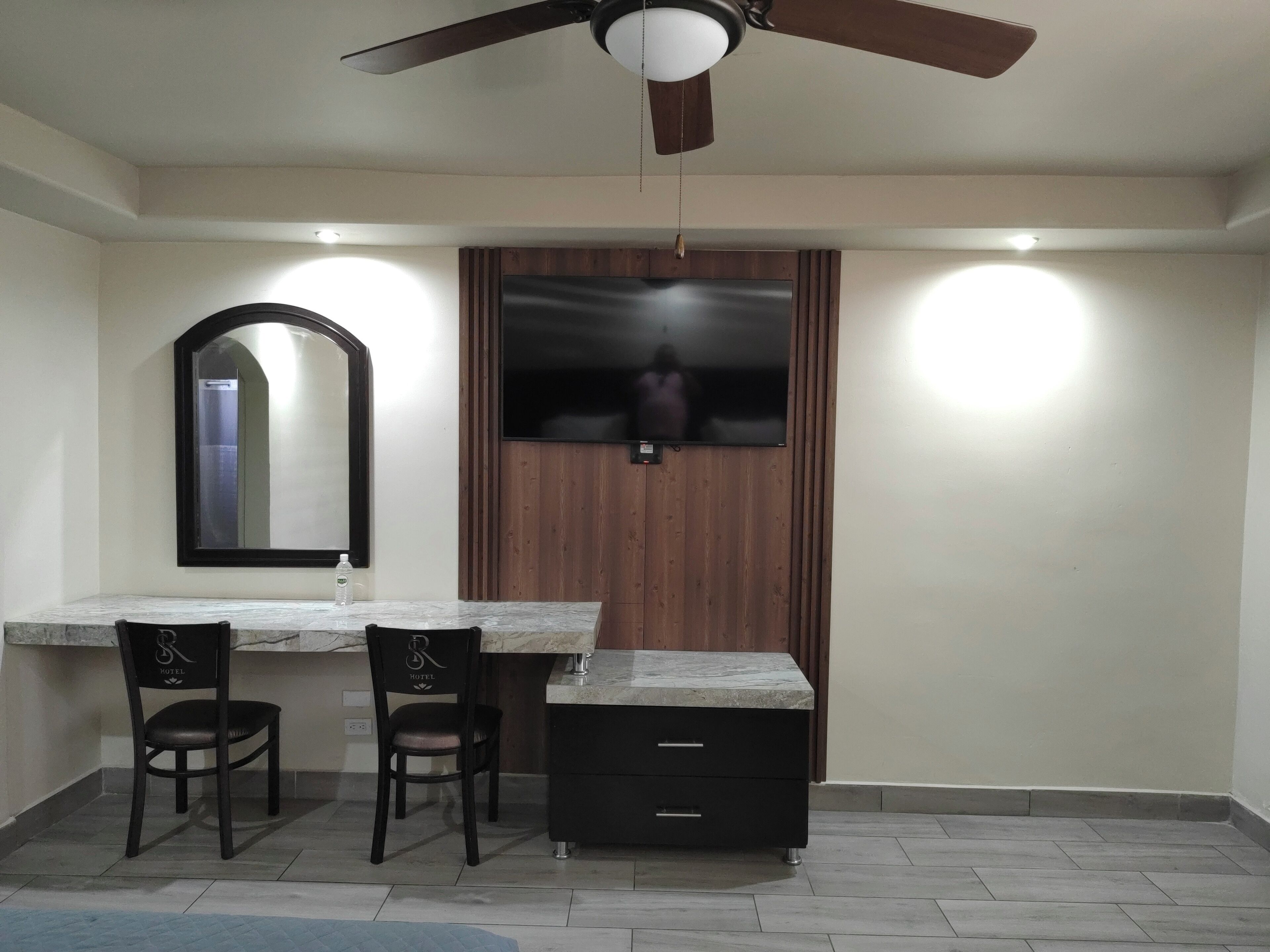 Quarto de hotel com escrivaninha, duas cadeiras, espelho, televisão, ventoinha no teto e parede revestida de madeira.