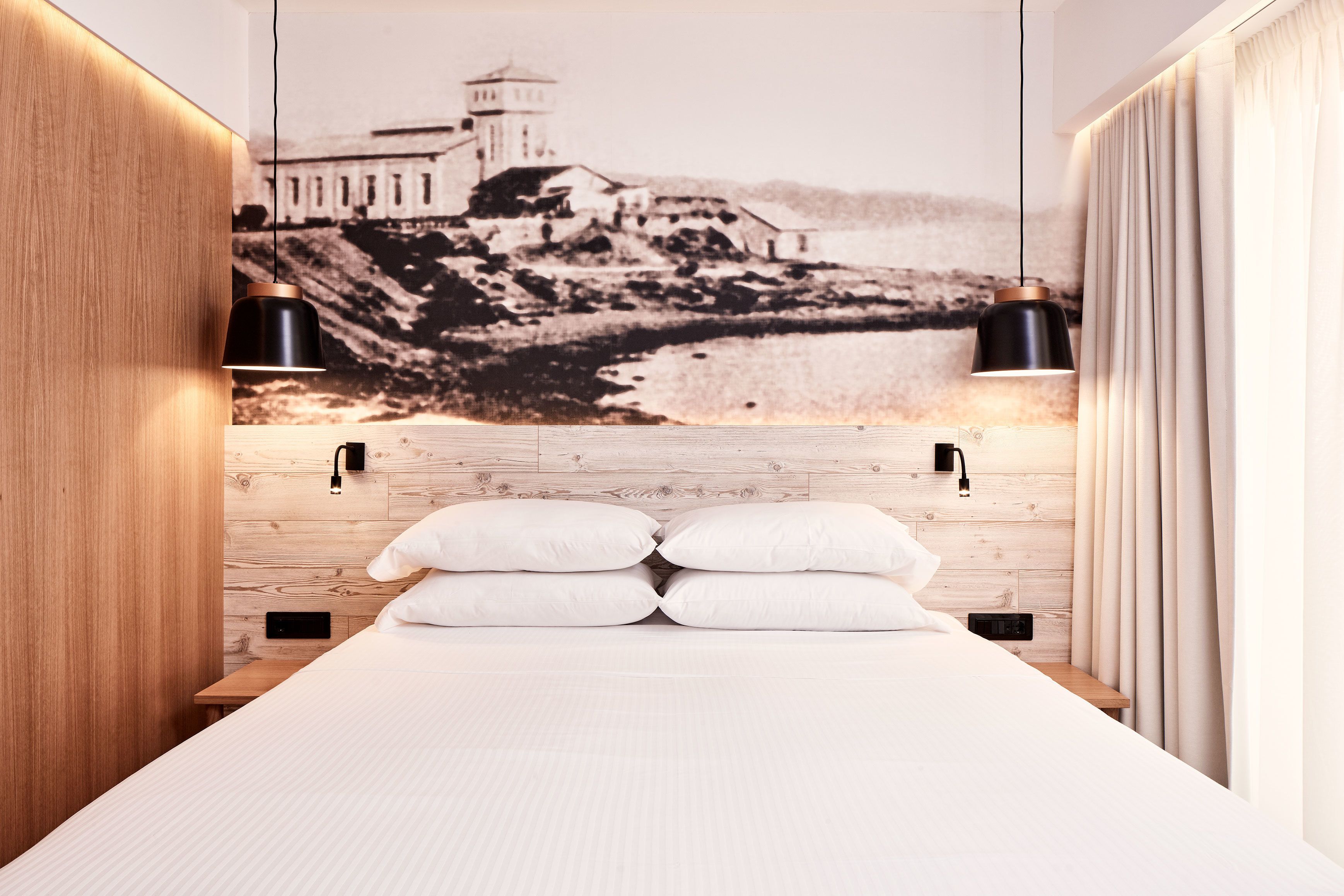 Quarto de hotel moderno com uma cama grande, duas luminárias de cabeceira, um painel de madeira na cabeceira e uma obra de arte pendurada na parede retratando um edifício histórico.