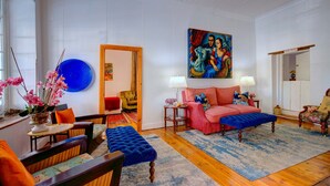 Uma sala de estar com um sofá vermelho, um banquinho azul e um quadro sobre o sofá. Há um prato azul na parede e um vaso com flores em uma mesa.