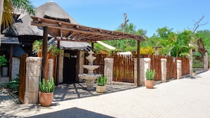 Entrada de um resort tropical com um edifício de telhado de palha, pérgola de madeira e cactos em vasos.