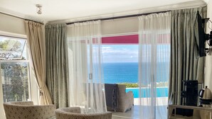 Quarto com duas poltronas estampadas, máquina de café e vista para o oceano através de cortinas translúcidas.
