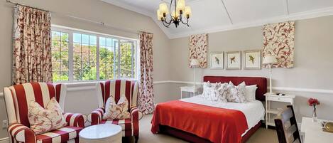 Quarto de hotel com uma cama grande, uma poltrona vermelha estofada, uma mesa redonda e uma janela com cortinas.