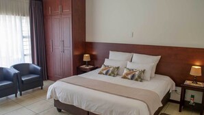 Quarto de hotel com uma cama grande, duas mesas de cabeceira, um guarda-roupa, uma cadeira e uma janela com cortinas.