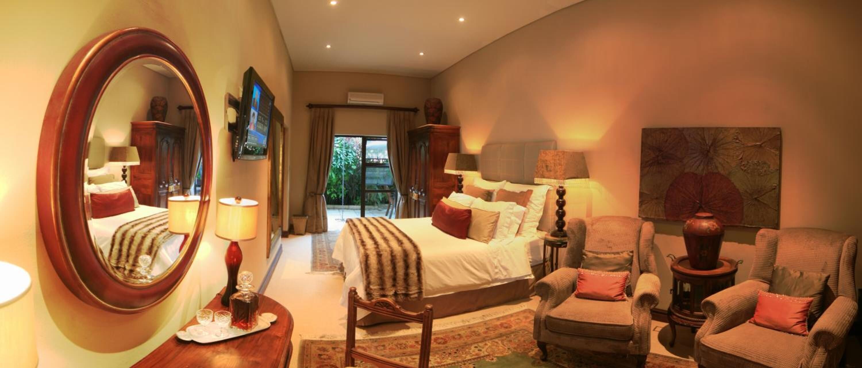 Quarto de hotel com uma cama grande, duas poltronas, uma mesinha de madeira, um espelho e um quadro na parede.