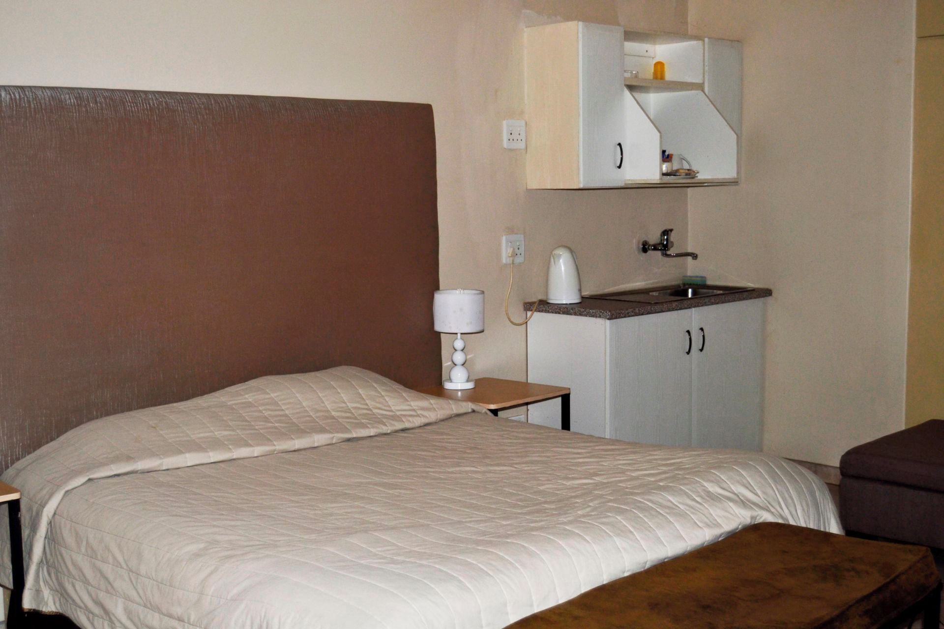 Quarto de hotel com cama, mesa de cabeceira, luminária, móvel pequeno com pia e uma cadeira.
