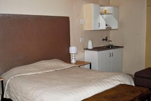 Quarto de hotel com cama, mesa de cabeceira, luminária, móvel pequeno com pia e uma cadeira.