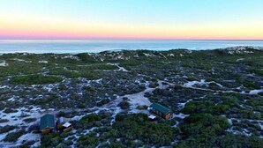 Vista aérea de uma área costeira com duas pequenas cabanas cercadas por vegetação densa e uma trilha de terra.